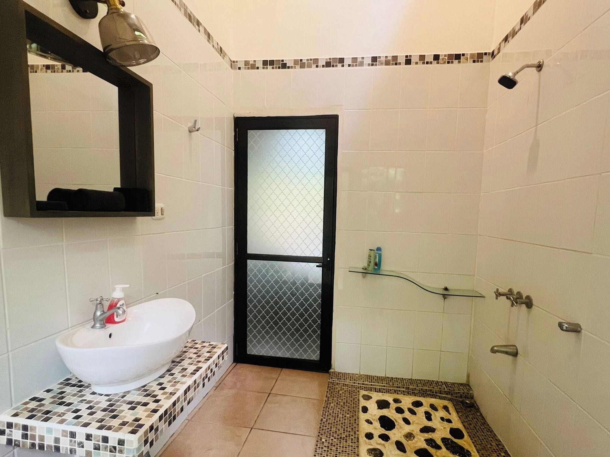 Casa de banho com lavatório branco, espelho, zona de duche e chão em azulejos.