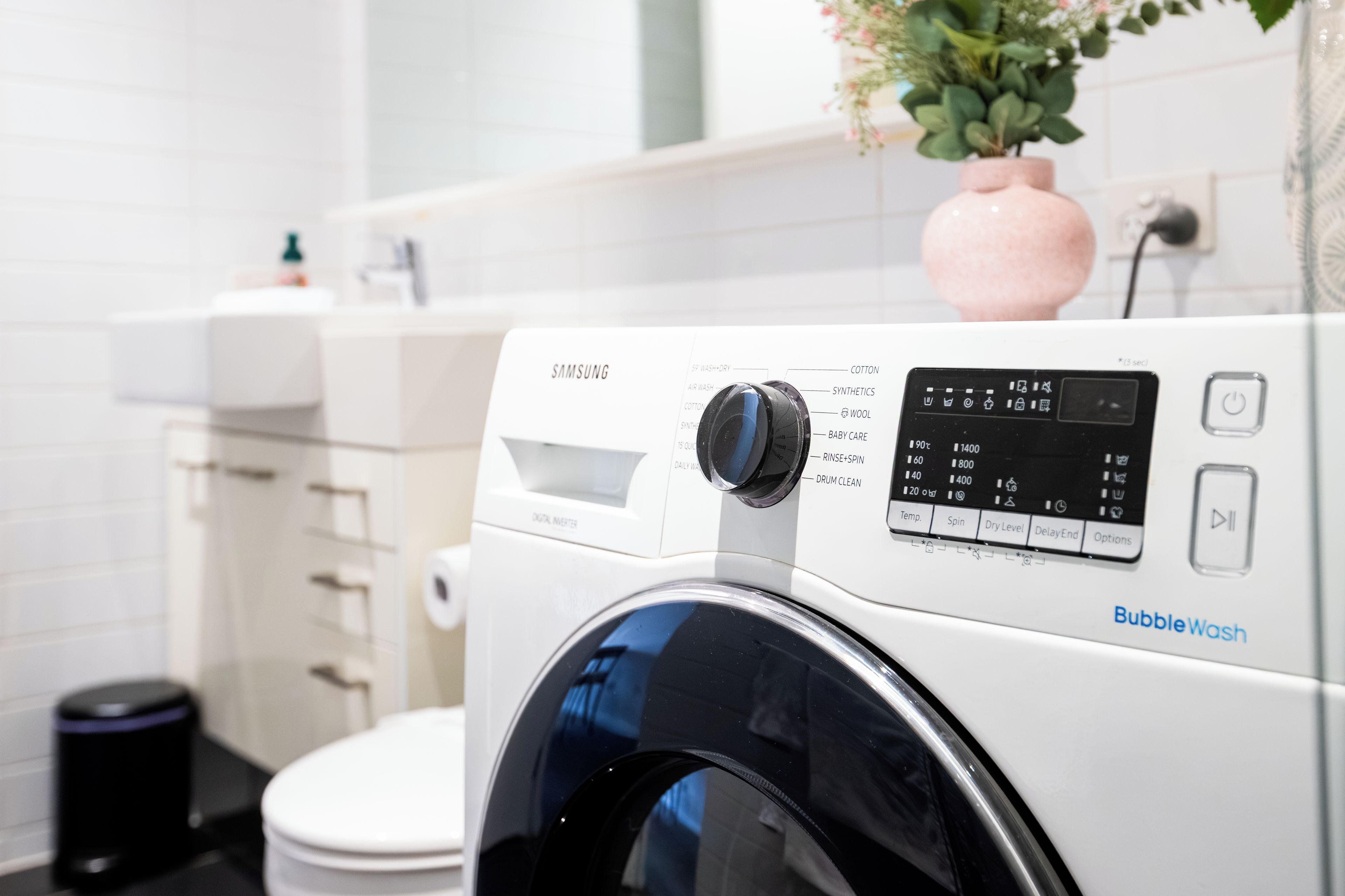 Máquina de lavar Samsung com interface digital, pia branca e um cesto de lixo em um banheiro.