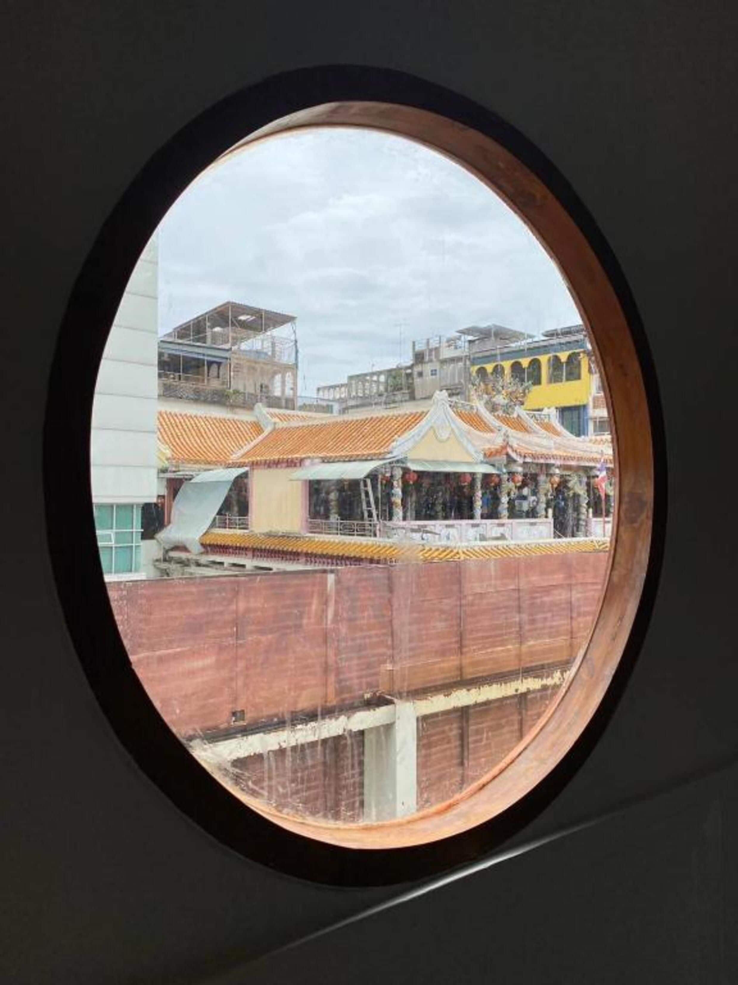 Visão através de uma janela circular de um edifício com arquitetura tradicional, incluindo telhado com azulejos e um pátio.