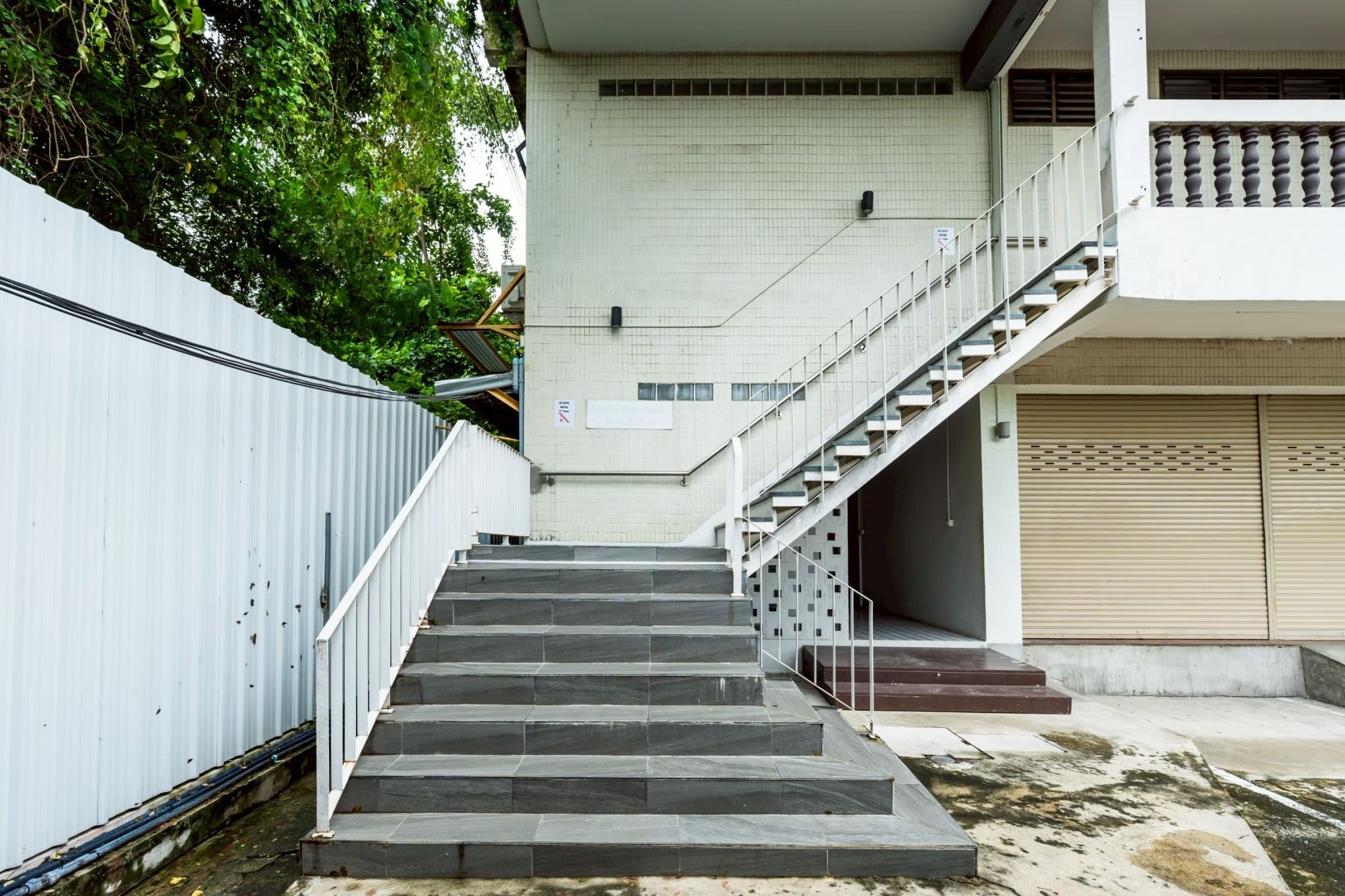 Escadas externas levam a um acesso de edifício, cercadas por uma grade de metal e vegetação.
