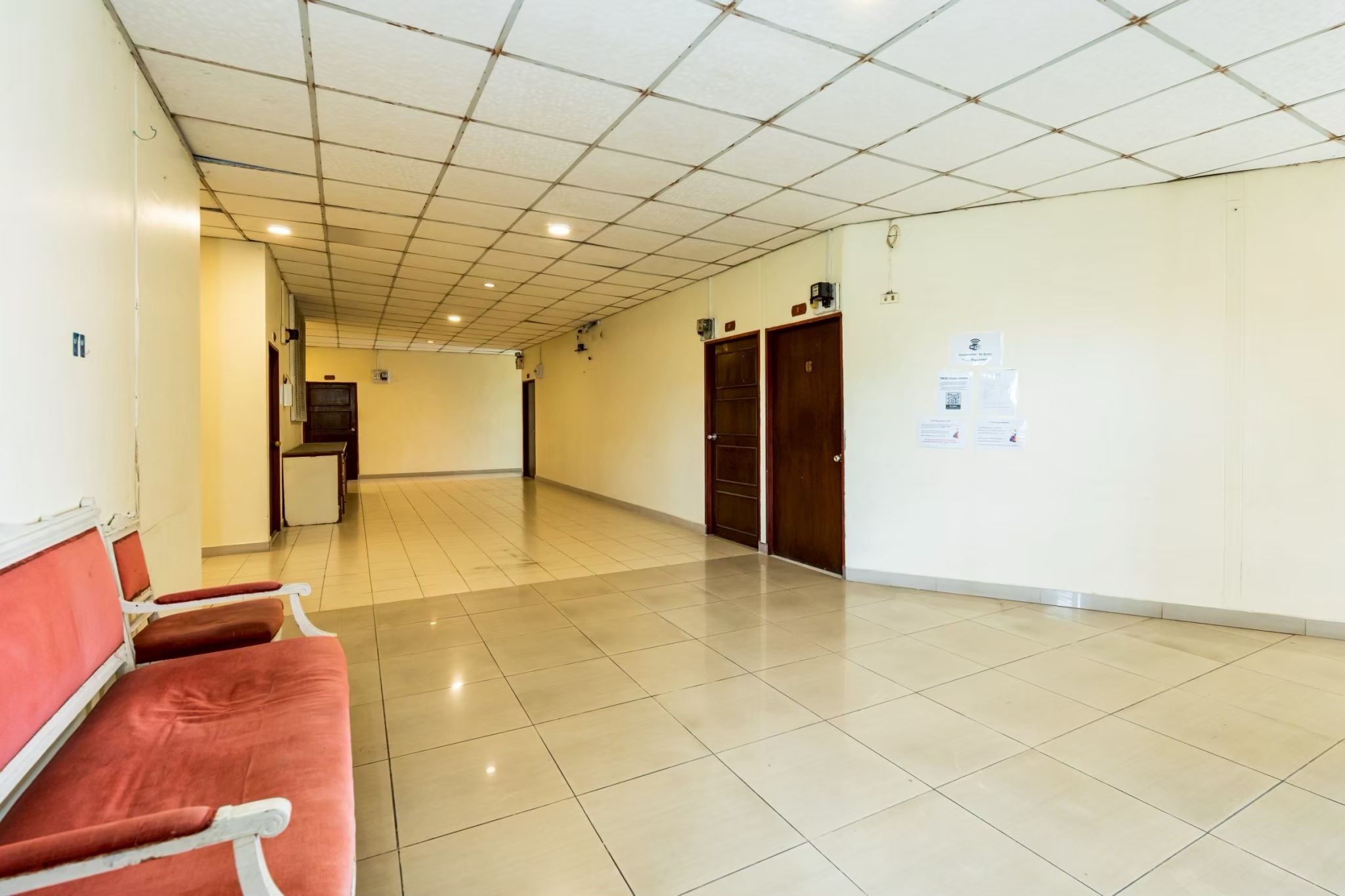 Corredor com piso ladrilhado, assentos vermelhos e portas em um dos lados.