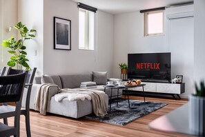 Uma sala de estar moderna com um sofá cinzento, uma mesa de centro e uma TV exibindo 'NETFLIX'.
