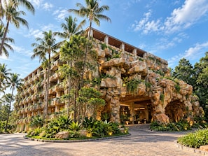 Um hotel com várias andares, fachada de pedra, palmeiras e área de entrada pavimentada.