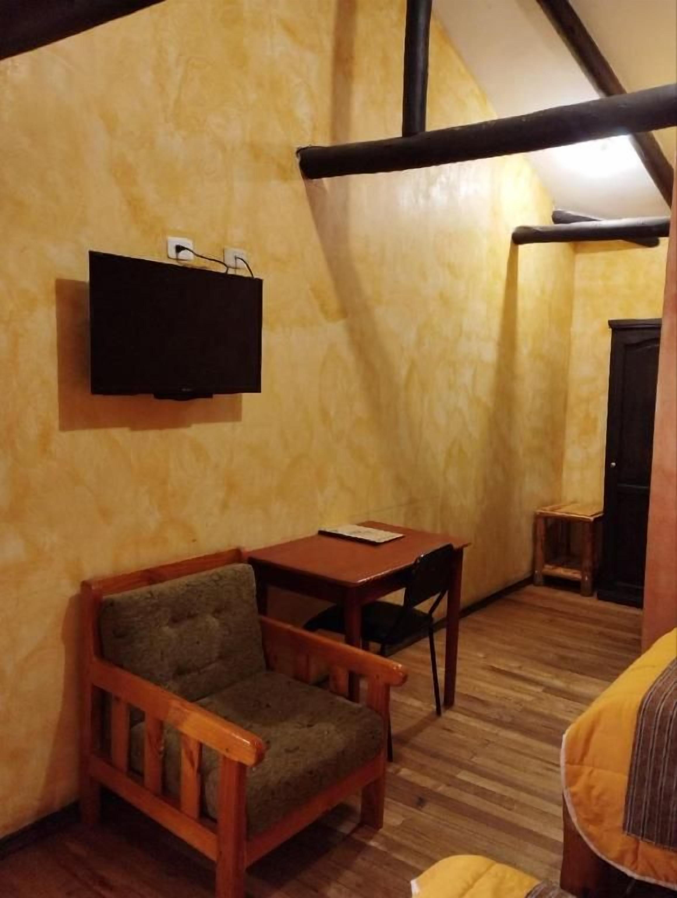 Quarto com cadeira de madeira, mesa pequena e televisão presa à parede.