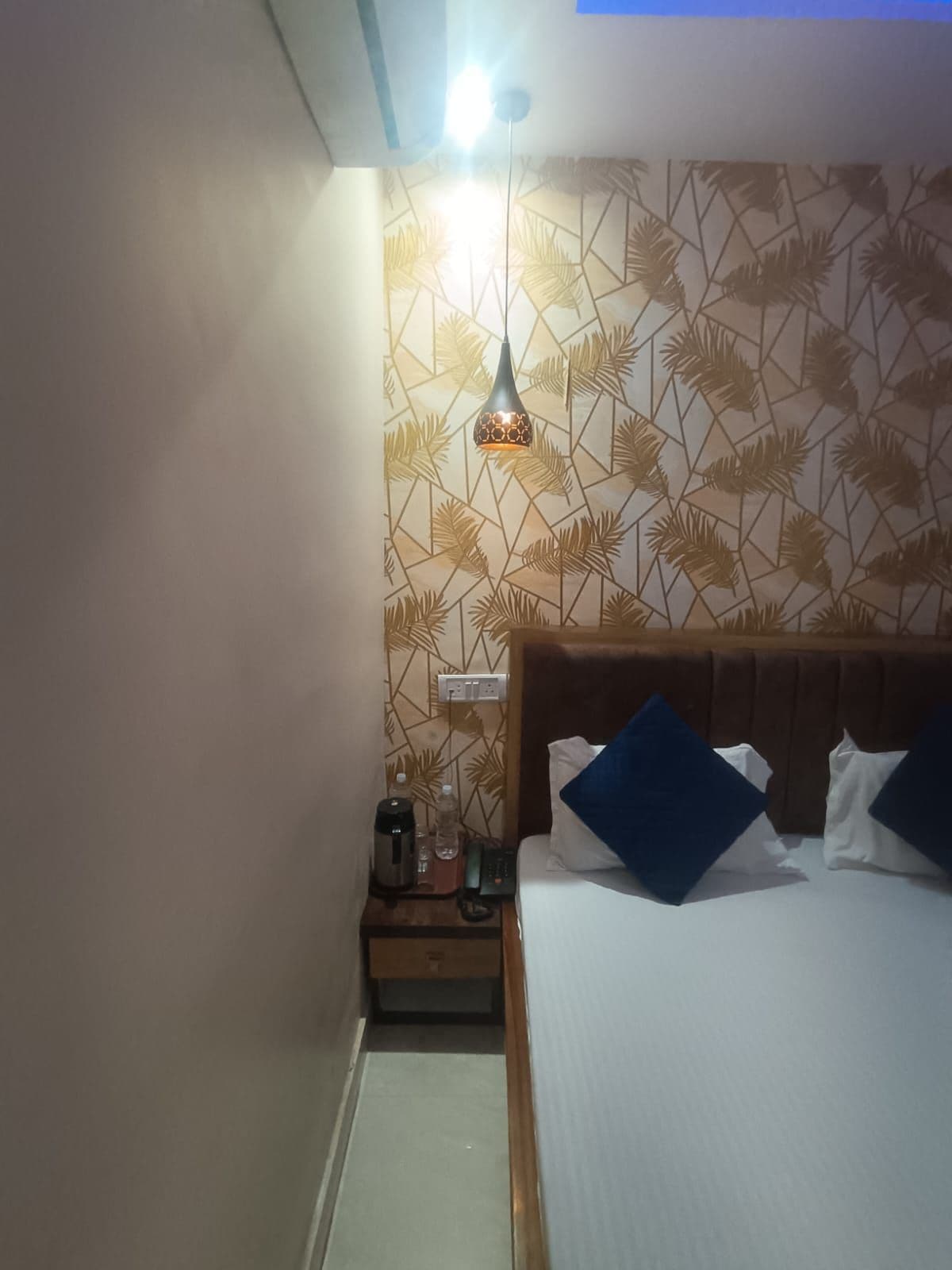 Quarto de hotel com cama, mesa de cabeceira, abajur e parede com padrão de folhas geométricas.