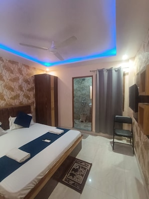 Quarto de hotel com cama, ventoinha no teto, área de chuveiro e televisão na parede.