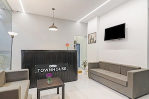 Uma área de recepção com um balcão preto identificado com 'OYO TOWNHOUSE', um sofá cinza, uma pequena mesa com um vaso, um lustre e uma televisão de tela plana fixada na parede.
