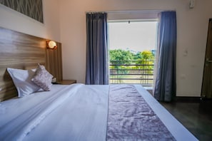 Quarto de hotel com cama, mesa de cabeceira, abajur e vista para o verde pela janela.