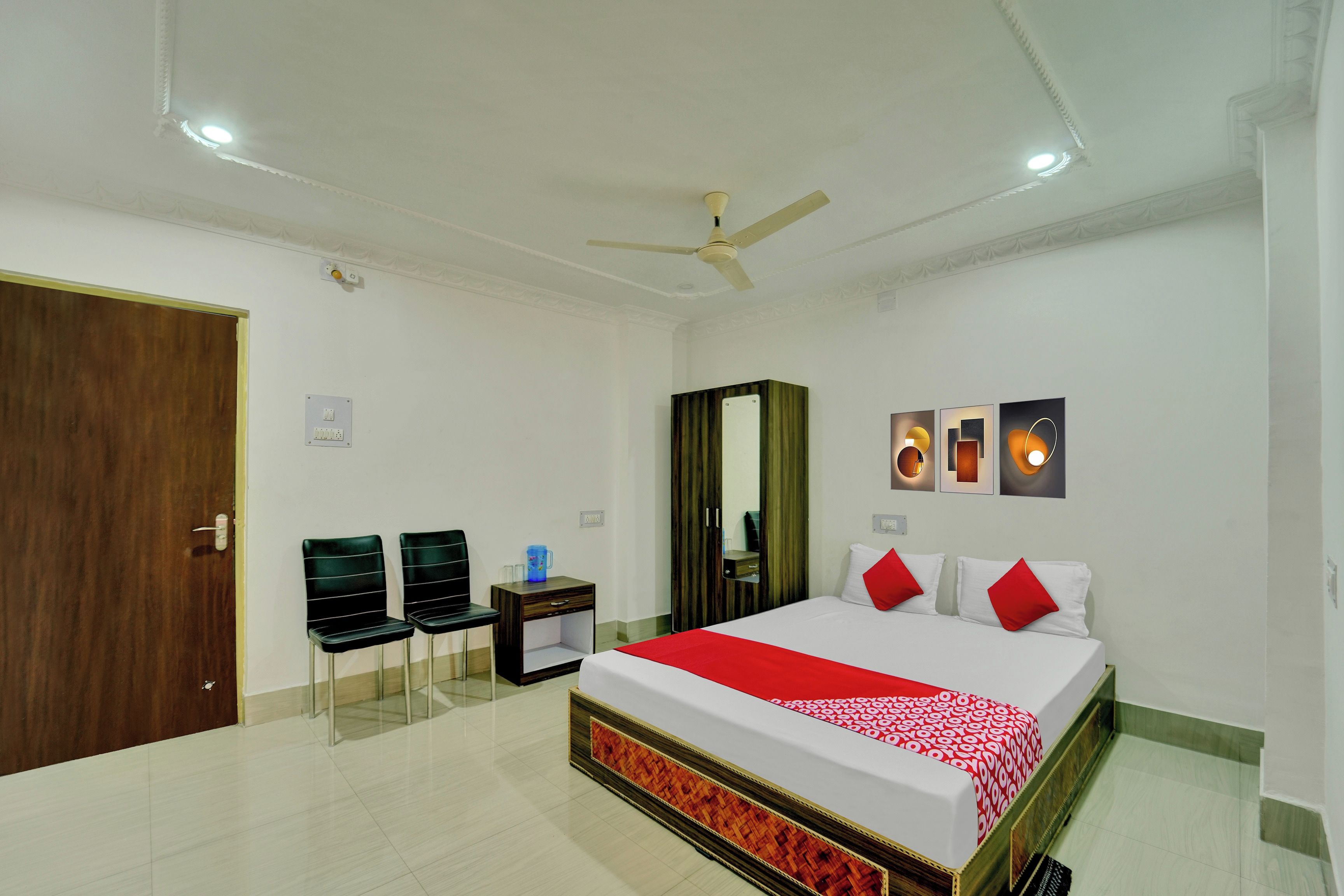 Quarto de hotel com cama, escrivaninha, duas cadeiras, guarda-roupa, ventoinha no teto e luzes na parede.