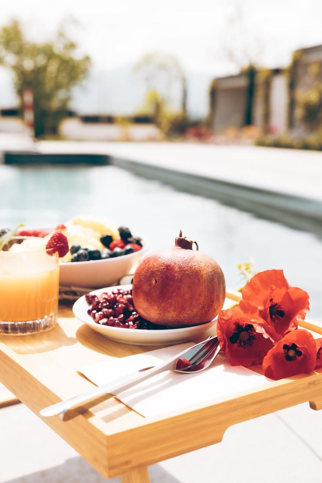 Bandeja de café da manhã com copo de suco de laranja, tigela de frutas vermelhas, romã e flores de papoula vermelha em mesa de madeira junto a uma piscina.