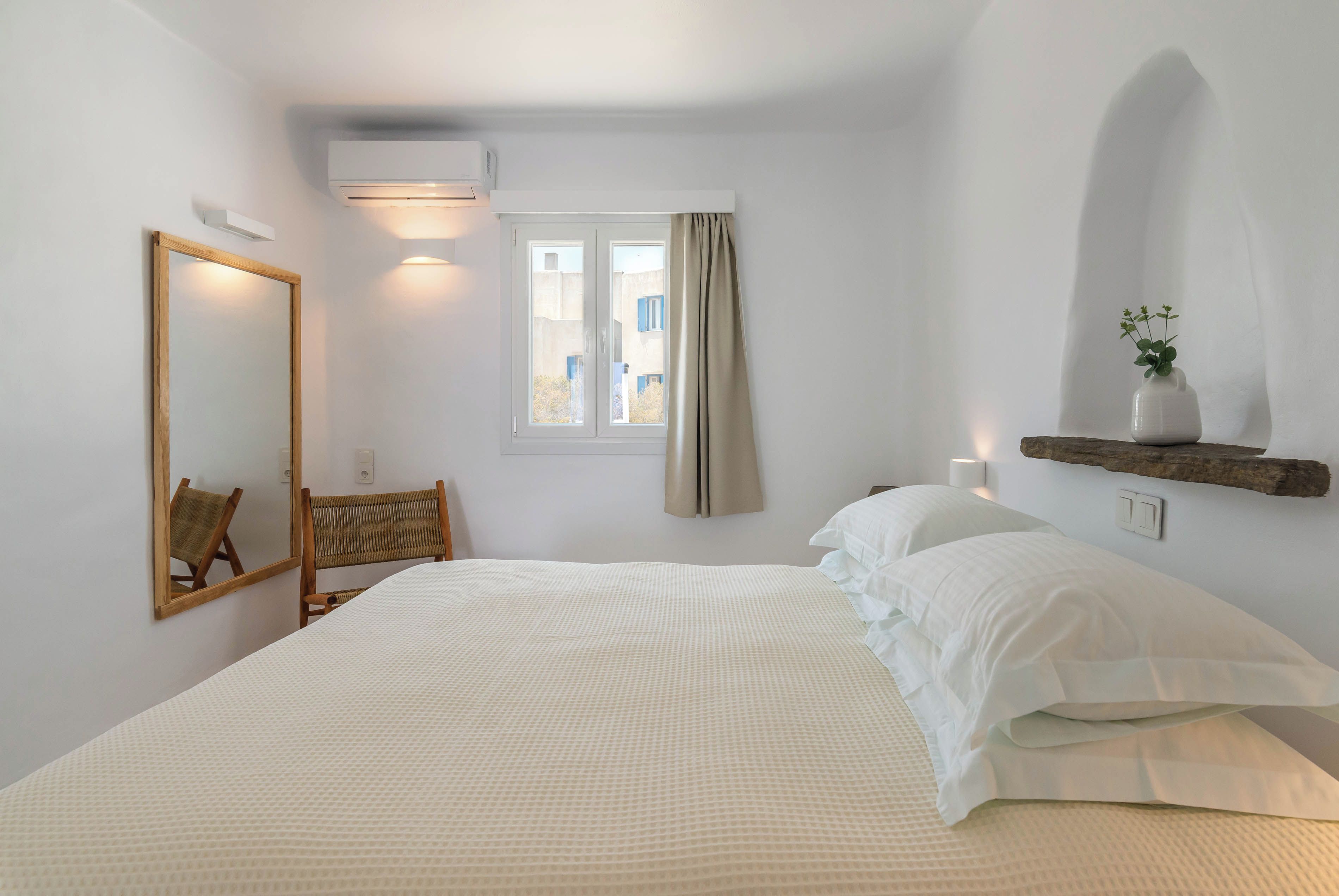 Quarto com cama, cadeira, espelho, janela com cortinas e prateleira com uma planta.