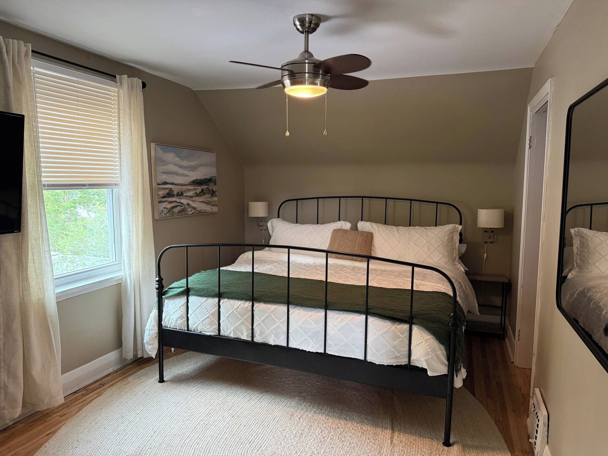 Quarto com cama, ventilador de teto, janela com cortinas, um quadro na parede e um espelho.