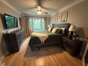 Quarto com cama, cômoda, mesa de cabeceira, abajur, ventilador de teto e televisão.