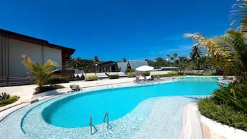 Piscina com borda curva, rodeada por palmeiras e um edifício com telhado plano.