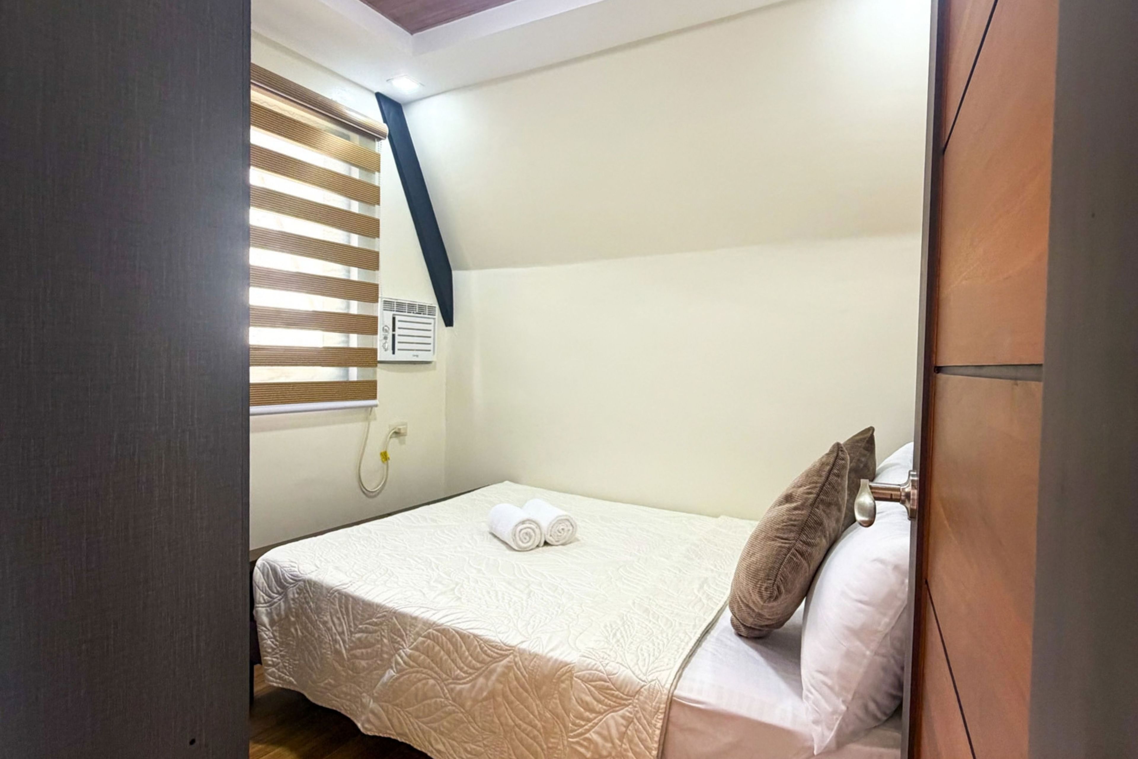 Quarto pequeno com uma cama de solteiro, guarda-roupa de madeira, janela com persianas e mesa de cabeceira com abajur.