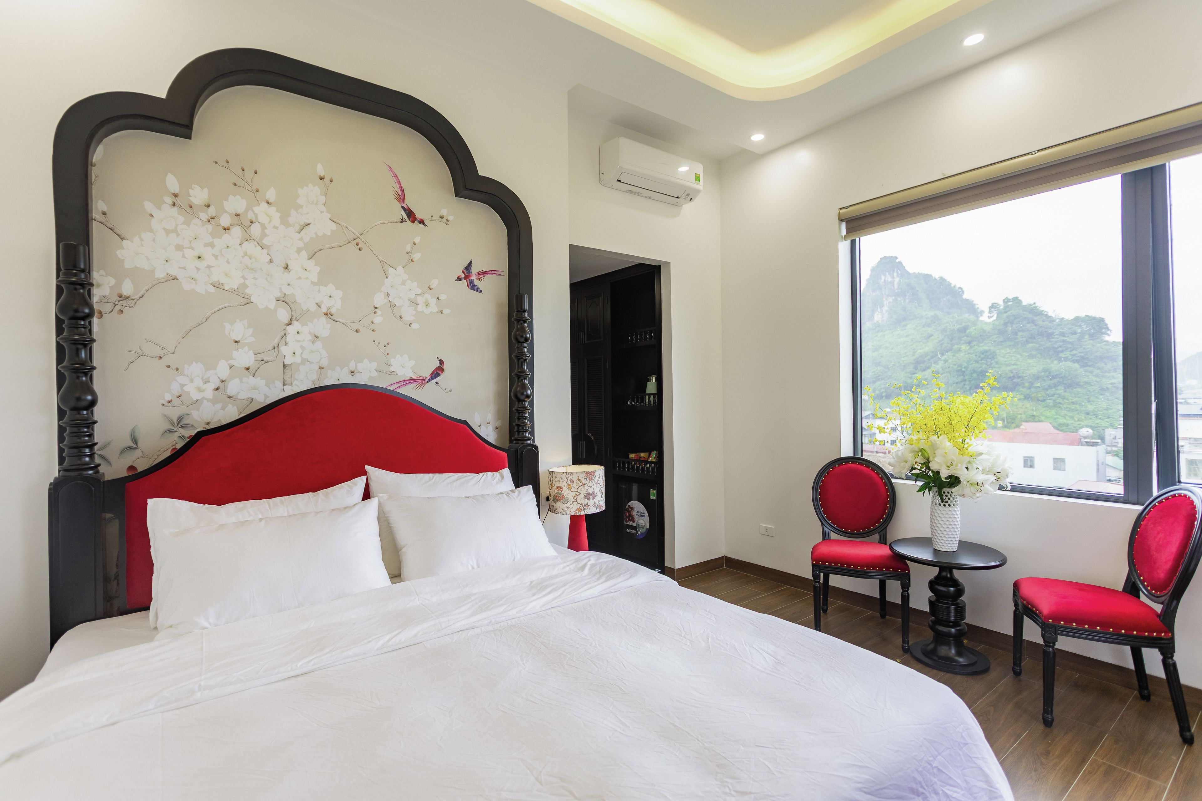 Quarto de hotel com uma cama grande, uma poltrona vermelha, uma mesinha de centro com um vaso, e uma janela com vista para áreas verdes e edifícios.