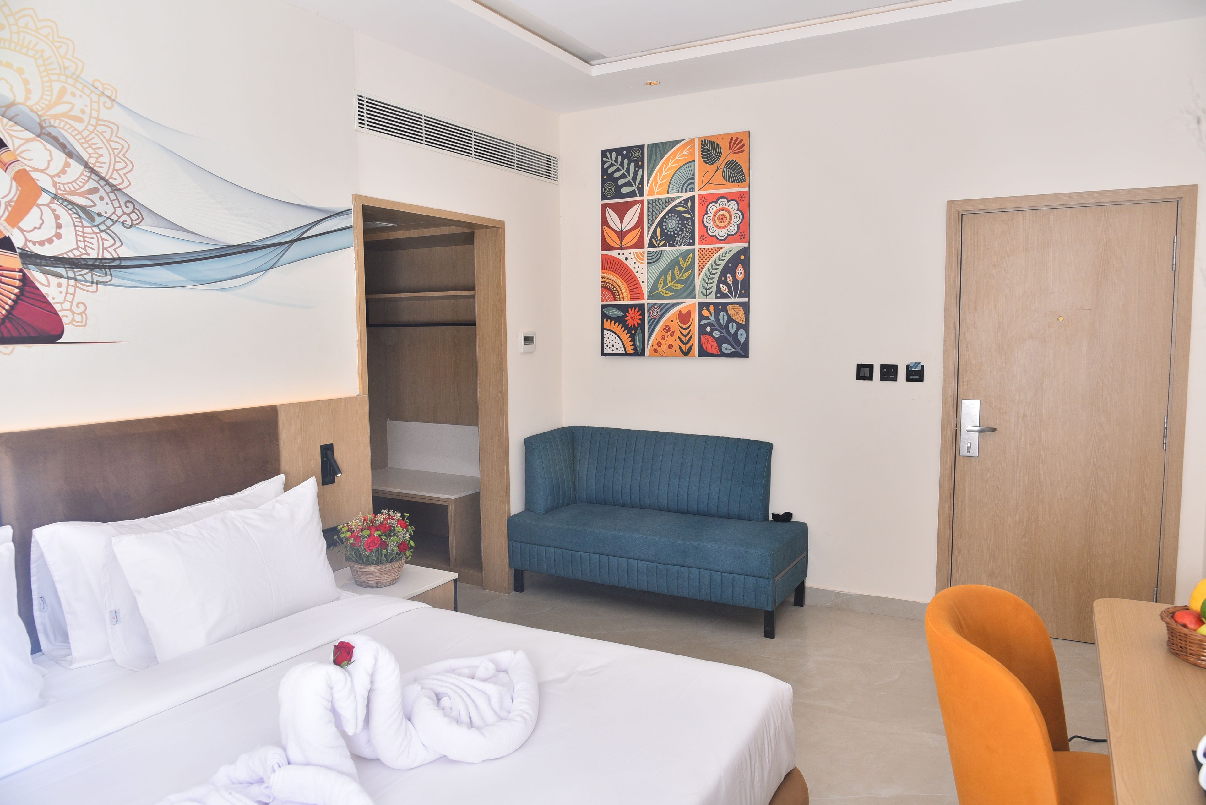 Quarto de hotel com cama, poltrona azul, armário de madeira, obra de arte na parede e uma porta.