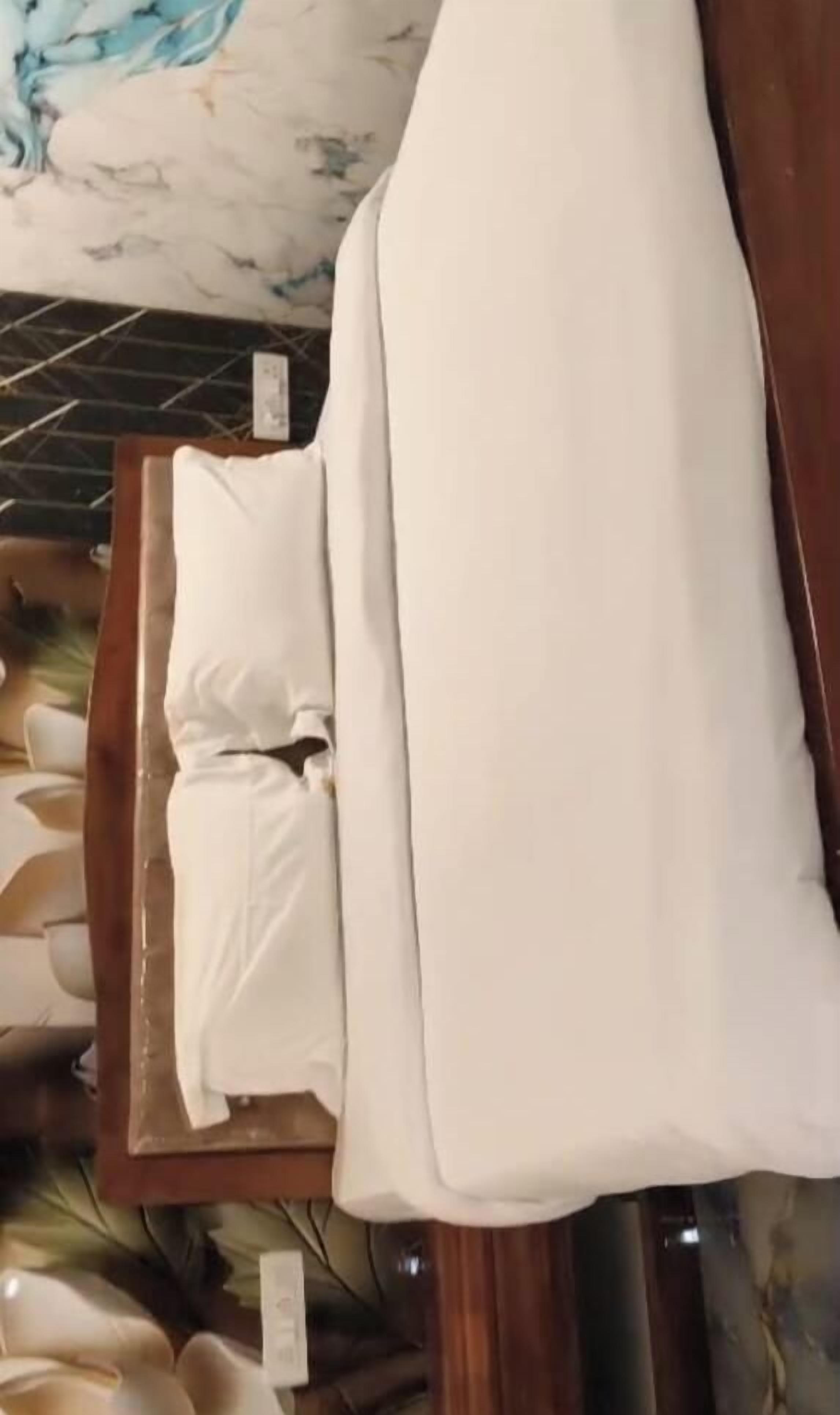 Cama bem arrumada com edredom branco e almofadas, cabeceira de madeira e parede com padrão marmorizado ao fundo.