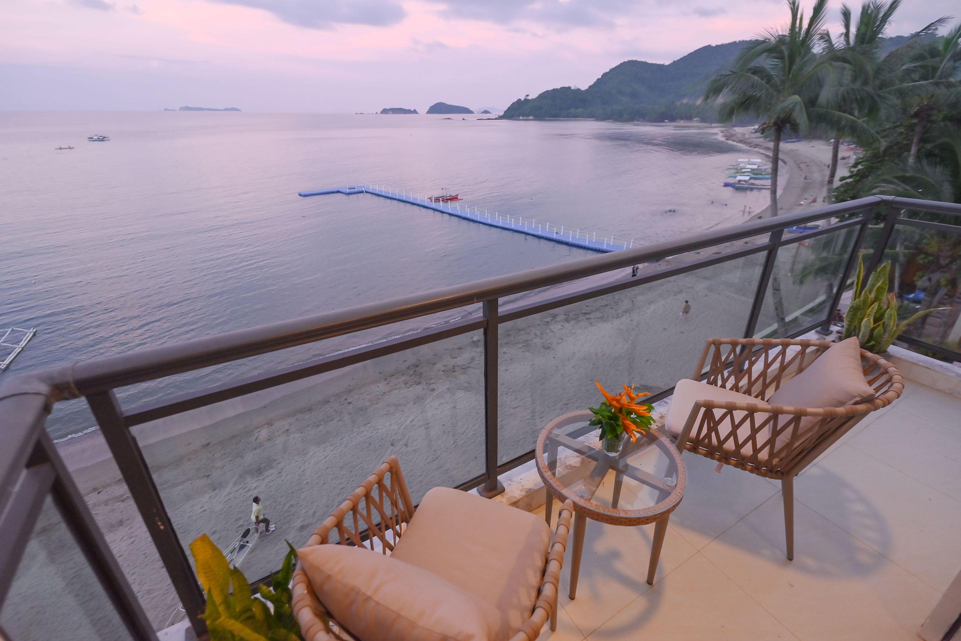 Varanda com móveis de vime, uma mesa pequena com um vaso de flores e vista para o mar, com barcos e um cais.