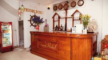 Bancada de recepção com acabamento em madeira e a inscrição 'Orient Hotel'. Há relógios na parede acima do balcão e uma geladeira Coca-Cola à esquerda.