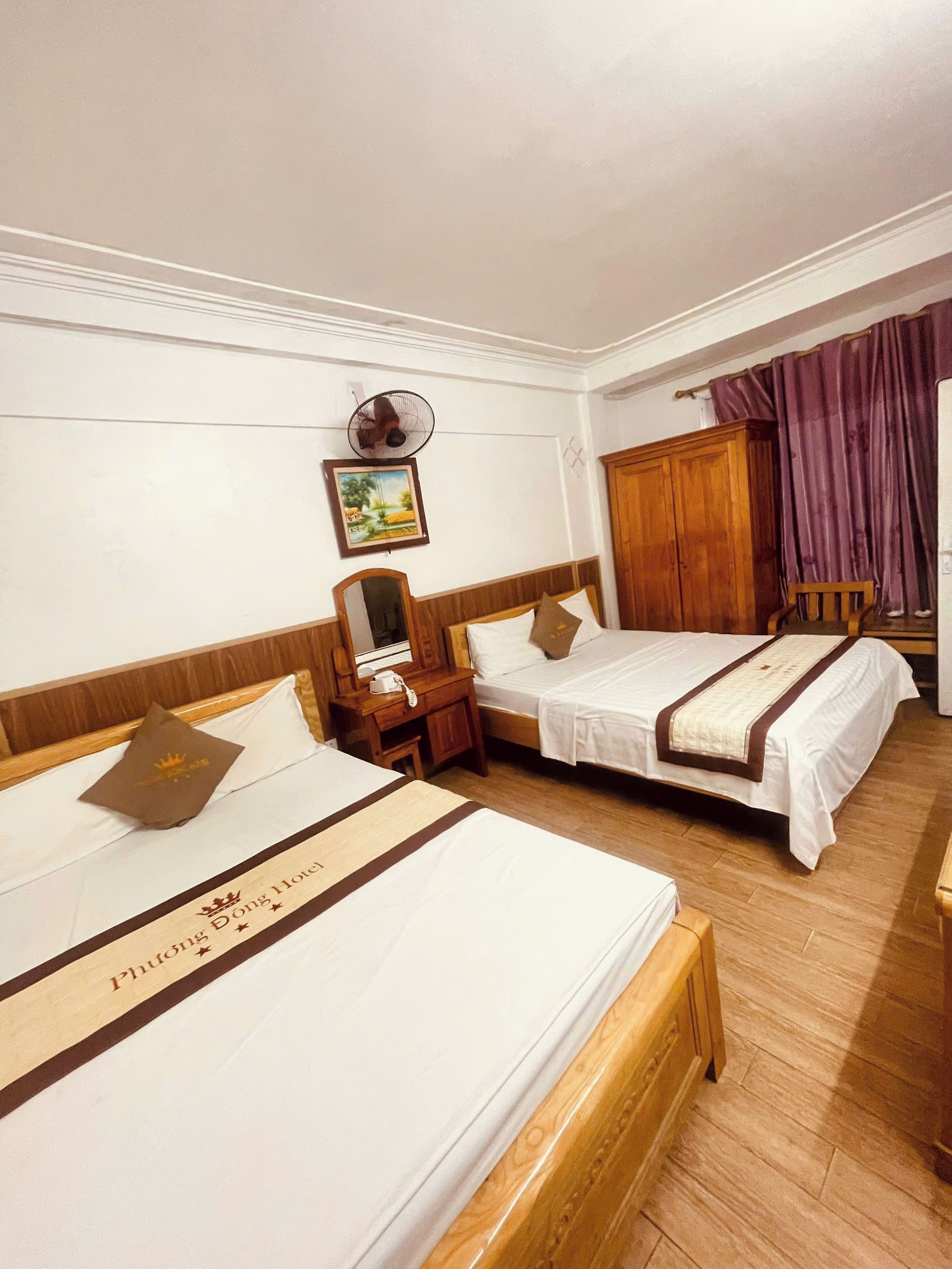 Quarto de hotel com duas camas de solteiro, um guarda-roupa de madeira, uma escrivaninha com telefone e um ventilador fixado na parede.