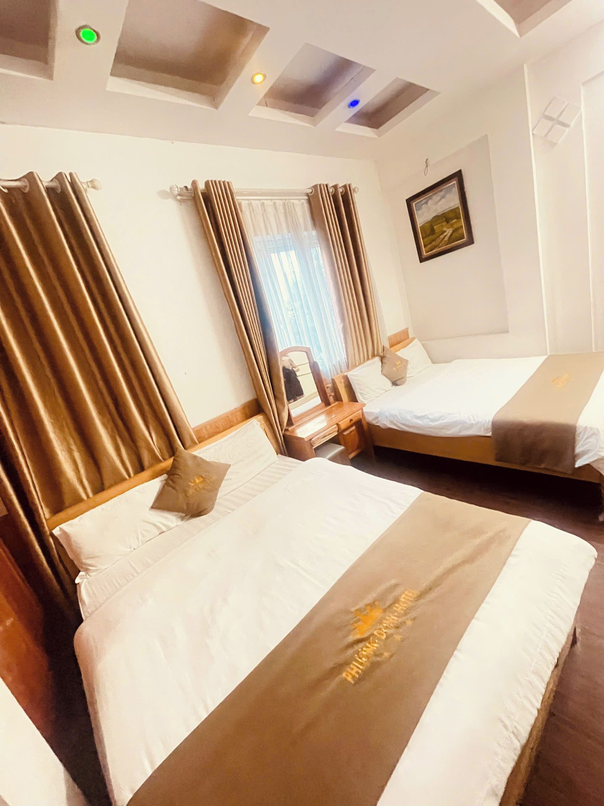 Quarto de hotel com duas camas, cabeceira de madeira, janela com cortinas, um quadro na parede e teto com claraboias.