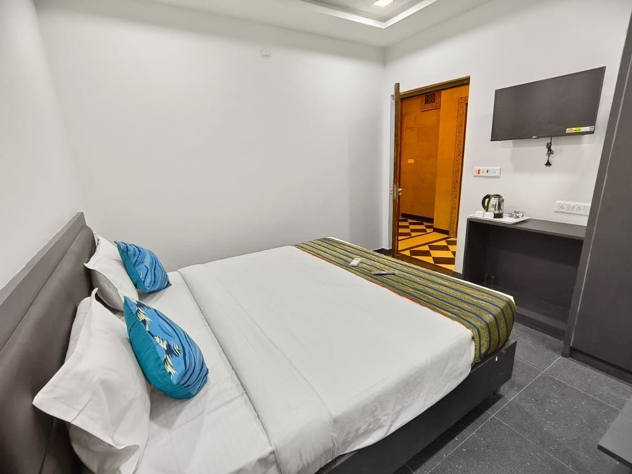 Quarto de hotel com cama, televisão presa na parede e uma pequena mesa com chaleira.