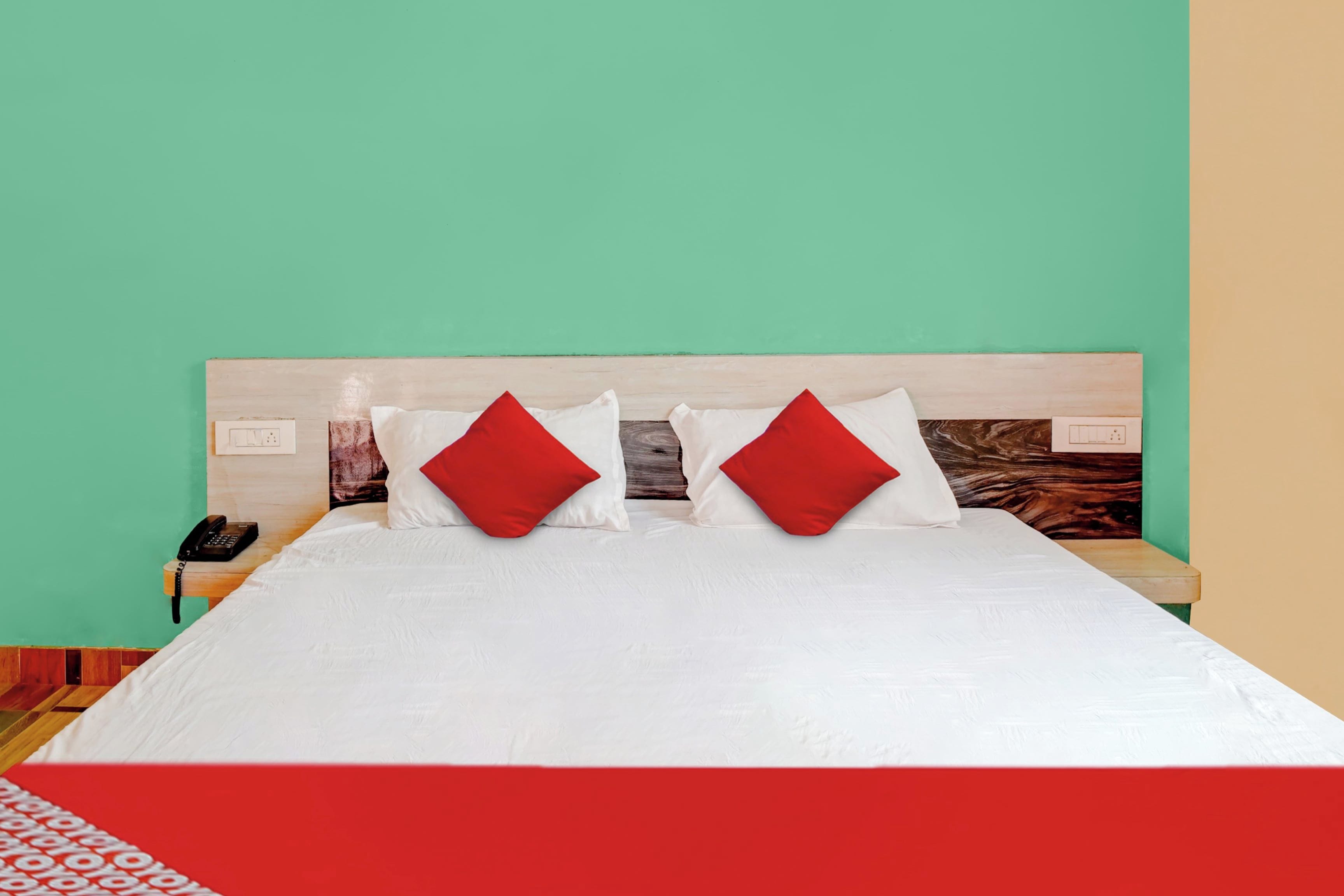 Cama com roupa de cama branca e duas almofadas vermelhas, cabeceira de madeira e mesa de cabeceira com um telefone.