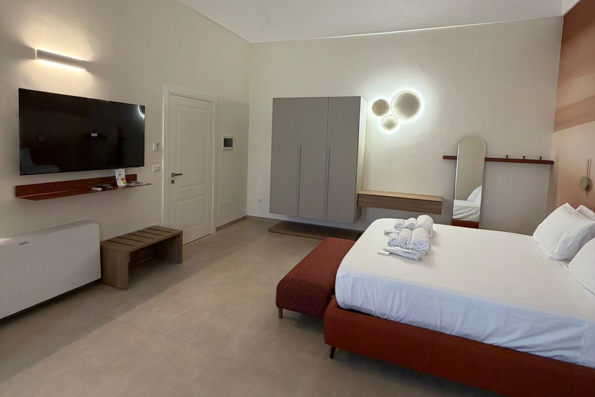 Quarto de hotel moderno com televisão de tela plana, guarda-roupa, cama com toalhas e um pequeno espaço de estar.