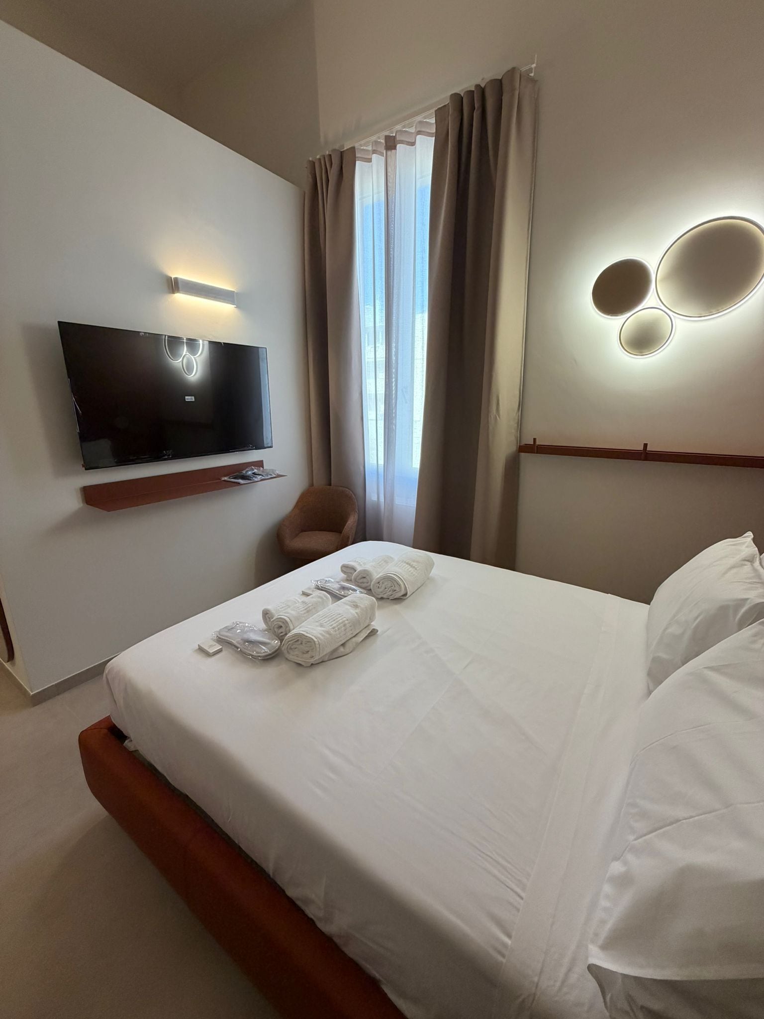 Quarto de hotel com cama, televisão na parede, uma cadeira e janela com cortinas.