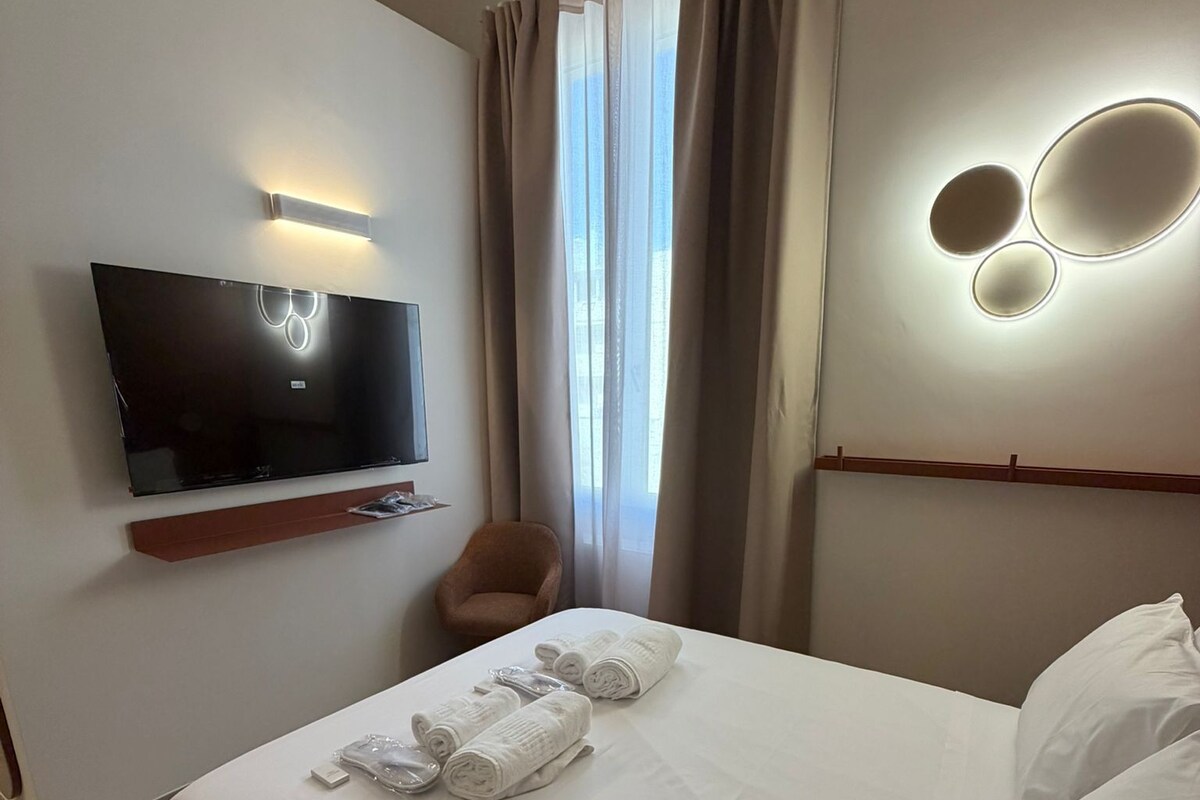Quarto de hotel com cama, televisão na parede, uma cadeira e janela com cortinas.