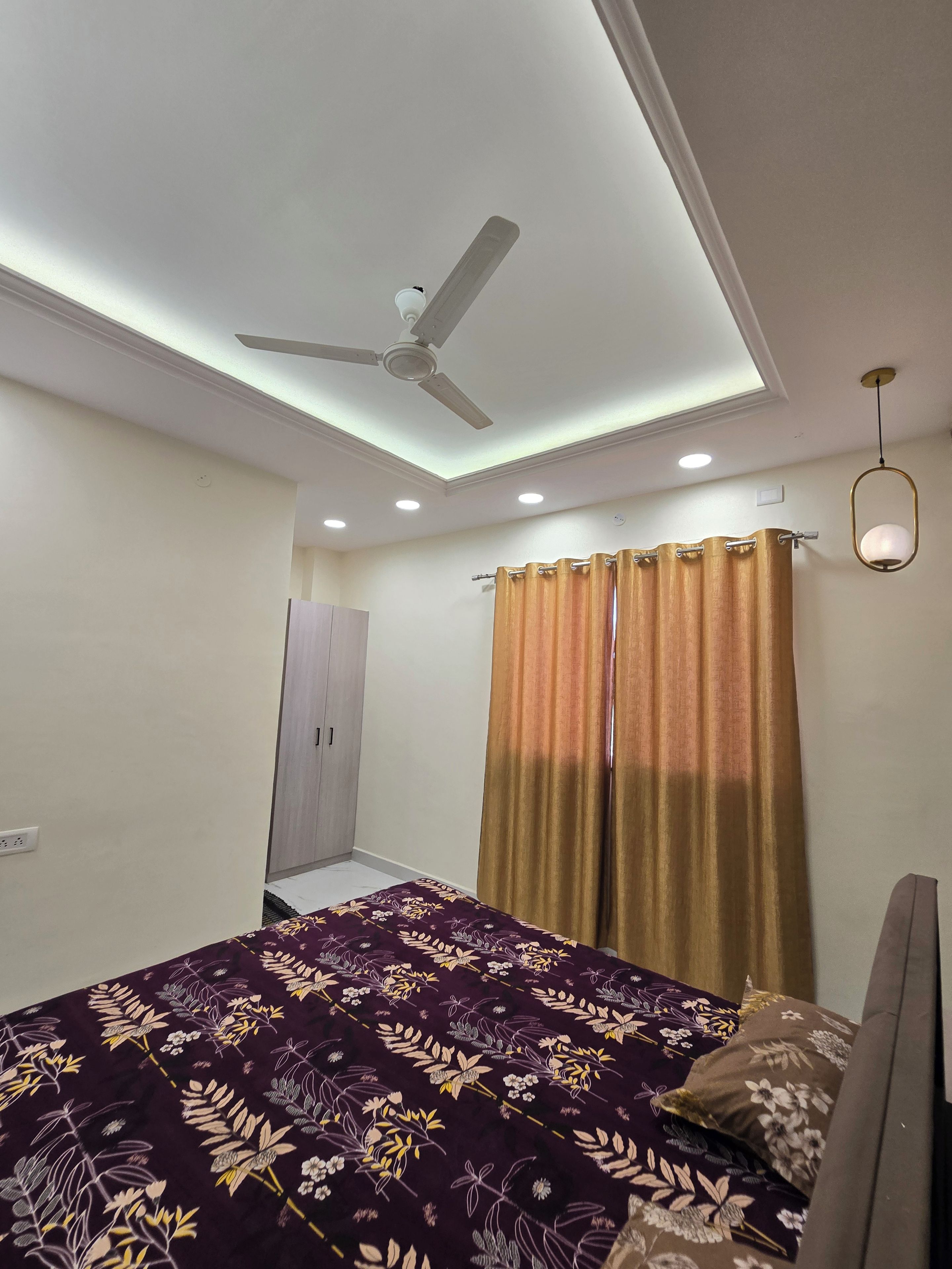 Quarto com cama, ventoinha no teto, iluminação embutida, guarda-roupa e janela com cortinas.