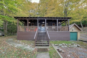Uma cabana com um deck de madeira, uma escadaria que leva a um alpendre coberto e uma porta de garagem verde.