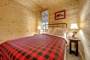 Quarto de cabana de madeira com uma cama, duas luminárias de cabeceira, uma janela com persianas e um quadro de urso na parede.