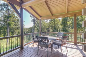 Um deck coberto ao ar livre com mesa e cadeiras, cercado por um corrimão de madeira, com vista para uma área arborizada.