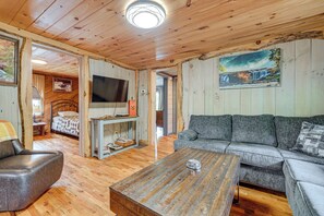 Interior acolhedor de uma cabana, com piso de madeira, um sofá, uma poltrona, uma televisão e um quadro na parede.