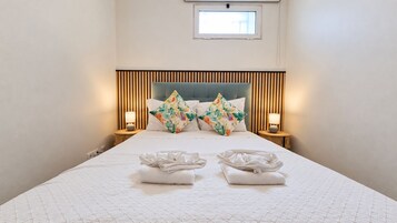 Quarto de hotel com cama, duas mesas de cabeceira, uma janela e cabeceira com ripas verticais.
