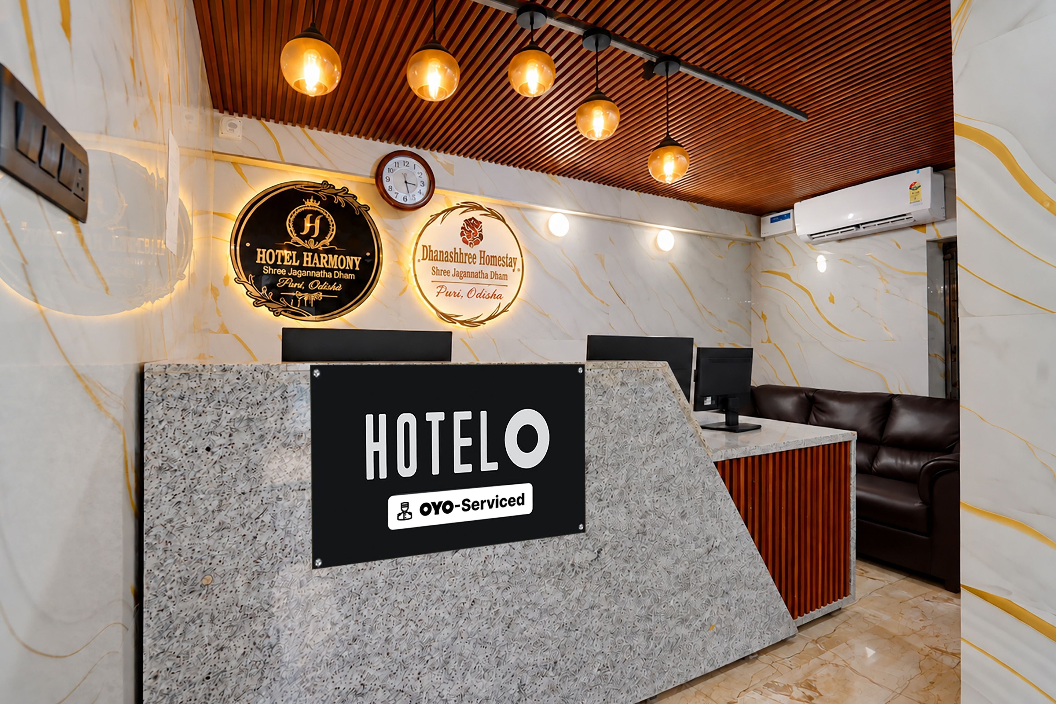 Balcão de recepção de hotel com placa indicando que é um serviço OYO. Existem duas placas acima do balcão, uma para Hotel Harmony e outra para Dhanashree Homestay, ambos localizados em Puri, Odisha.
