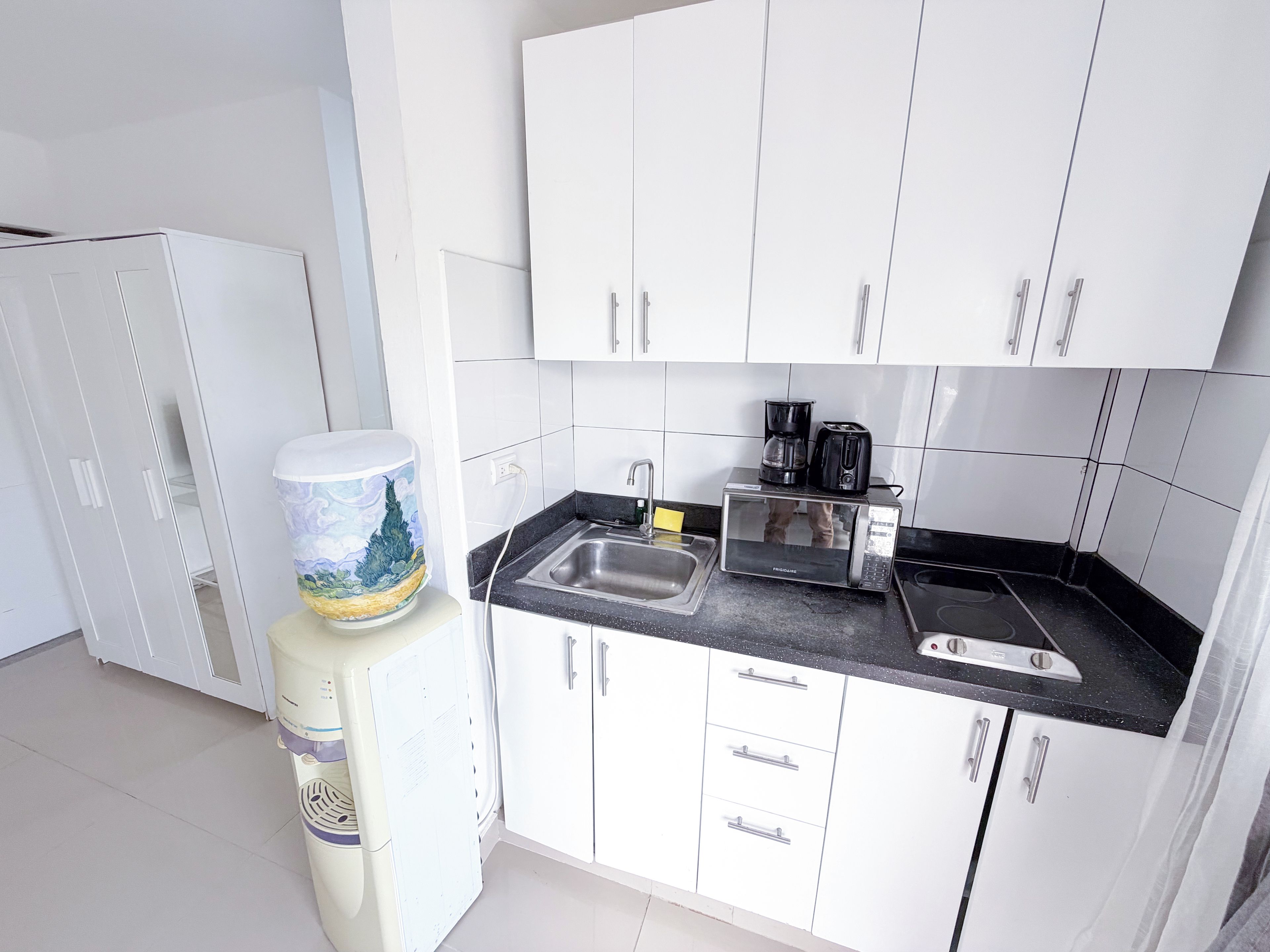 Uma cozinha moderna com armários brancos, bancada preta, pia, máquina de café e dispensador de água.