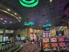 Interior de um cassino com máquinas de jogo, área de bar e uma grande tela.