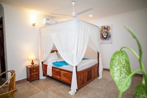 Quarto com cama de dossel, mesa de cabeceira com abajur, ventilador de teto e ar condicionado fixado na parede.