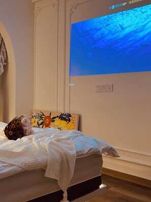 Cama com cobertor branco, buquê de flores e TV na parede exibindo uma cena subaquática azul.