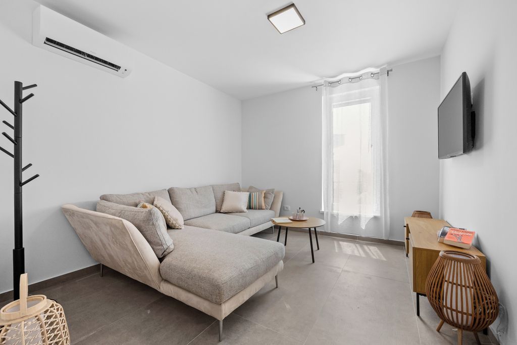Uma sala de estar moderna com sofá seccional, mesa redonda pequena, TV fixada na parede e janela com cortinas leves.