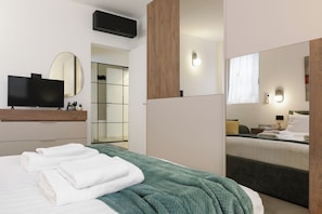 Quarto de hotel moderno com cama, televisão, espelho e banheiro com porta de vidro.