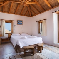 Quarto com cama, mesas de cabeceira, um banco, janela com cortinas, um quadro e vista para o mar.
