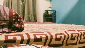 Quarto com uma cama, uma mesa redonda com diversos objetos, uma mesinha de cabeceira com abajur e uma janela com cortinas.