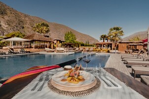Área de refeições à beira da piscina, com mesa posta para refeição, com vista para uma piscina com espreguiçadeiras e guarda-sóis.