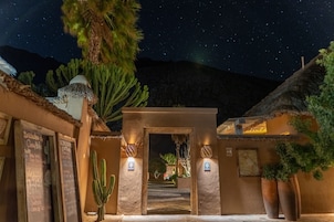 Vista noturna da entrada de um resort, com um caminho que leva a um edifício, cercado por cactos e palmeiras, sob um céu estrelado.