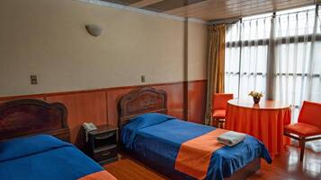 Quarto de hotel com duas camas, uma mesinha com cadeiras e uma janela com cortinas.