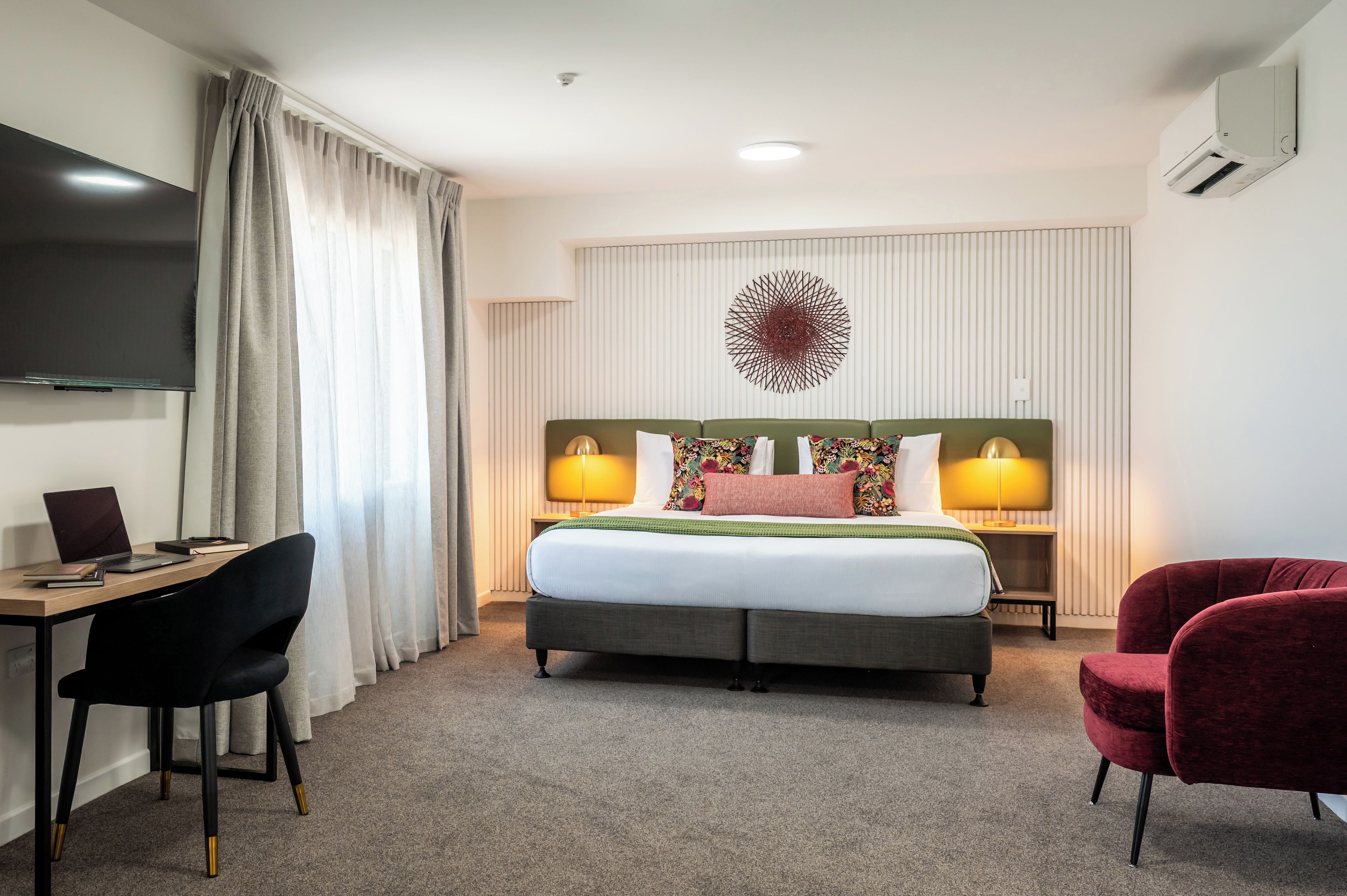 Quarto de hotel com cama, duas luminárias de cabeceira, escrivaninha com laptop, televisão e uma cadeira vermelha.